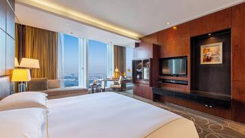 Quarto de hotel com uma cama grande, uma escrivaninha com cadeira, uma televisão na parede e vista para a cidade através de grandes janelas.