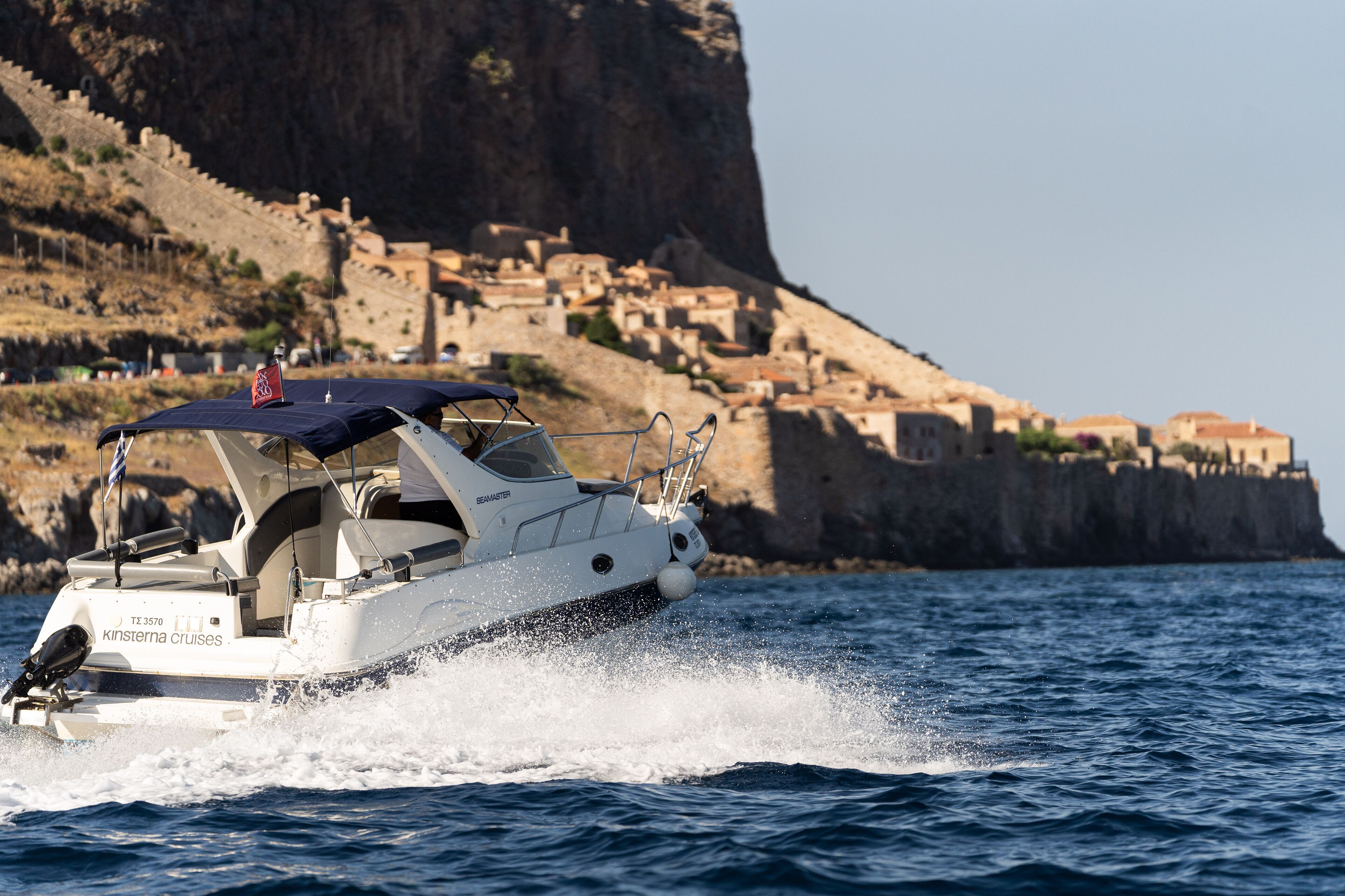 Um barco com toldo e a marcação "kinsterna cruises" na lateral, navegando pela água com um litoral rochoso e edifícios ao fundo.