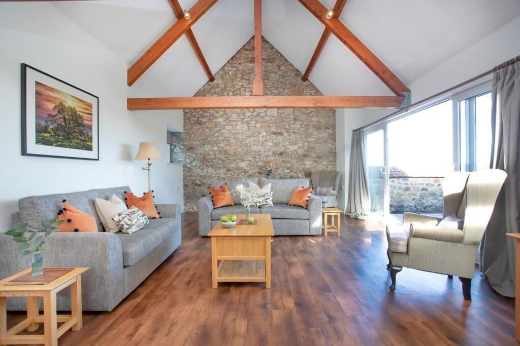 Uma sala de estar espaçosa com parede de pedra, vigas de madeira, um sofá cinzento, uma mesa de centro de madeira e uma poltrona confortável.