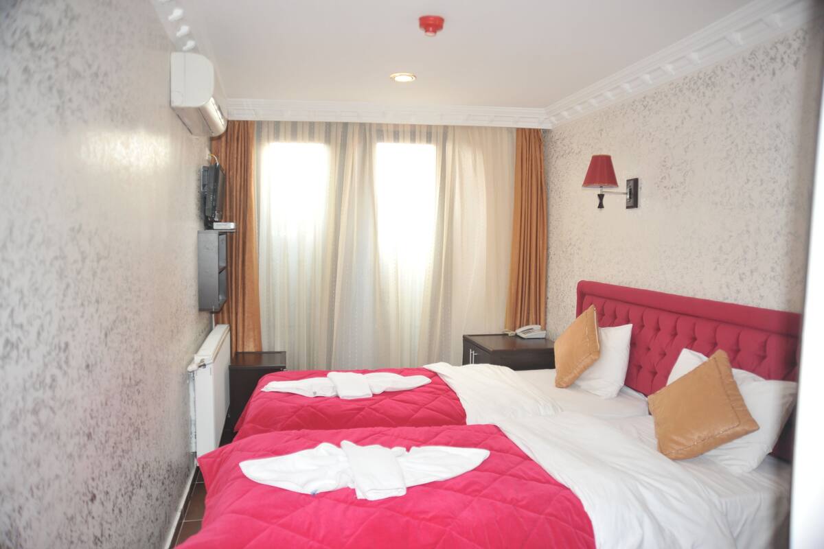 Quarto de hotel com duas camas, cabeceira vermelha, roupa de cama branca e janela com cortinas.