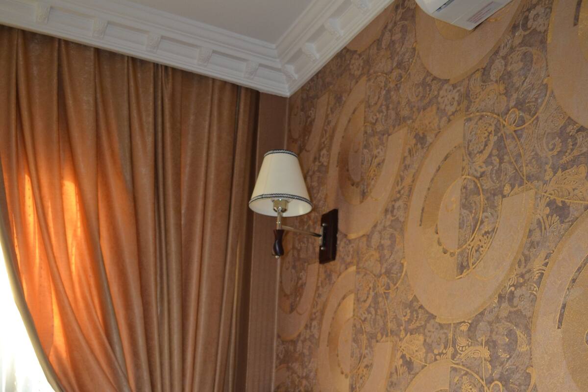 Quarto de hotel com cama, cortinas bege, luminária fixada na parede e papel de parede estampado.