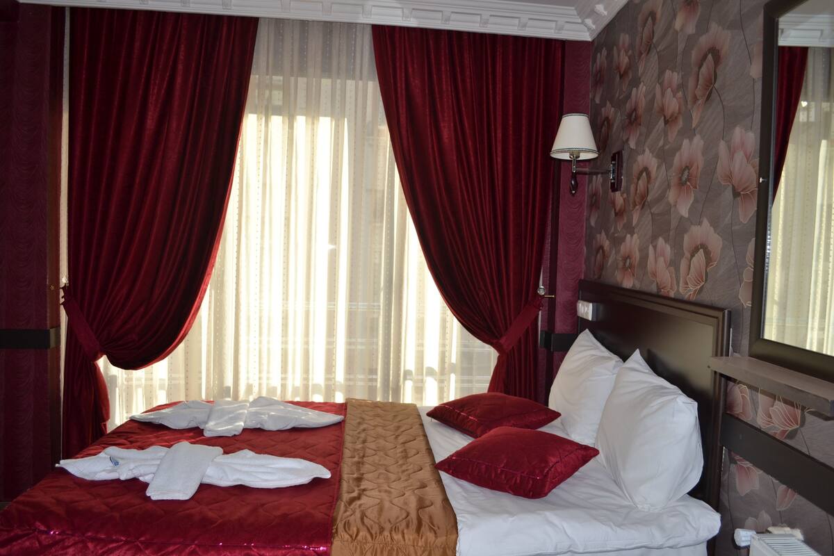 Quarto de hotel com cama, cortinas vermelhas, janela com cortinas translúcidas, luminária fixada na parede e papel de parede com padrão floral.