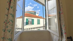 Quarto com janela que dá para um edifício com telhado de telhas vermelhas e persianas verdes.