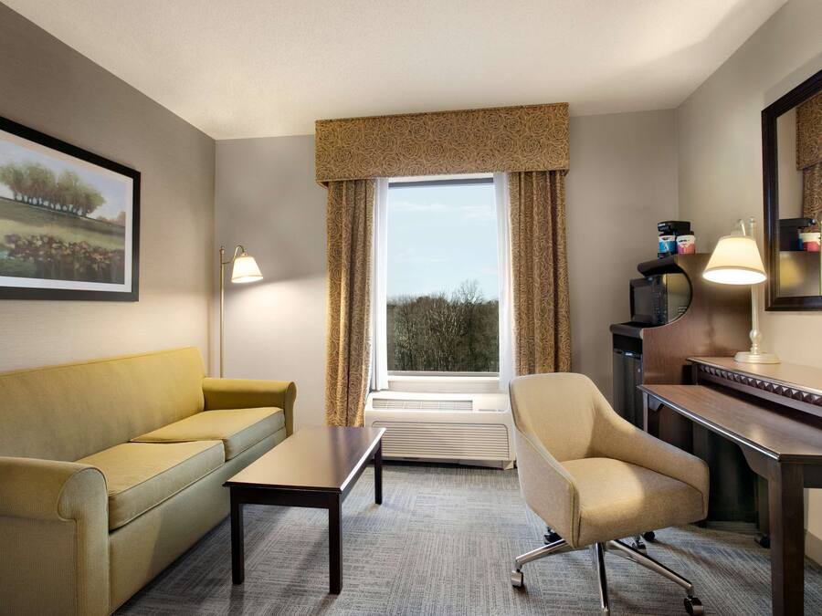 Quarto de hotel com sofá, poltrona, escrivaninha e uma grande janela com cortinas.