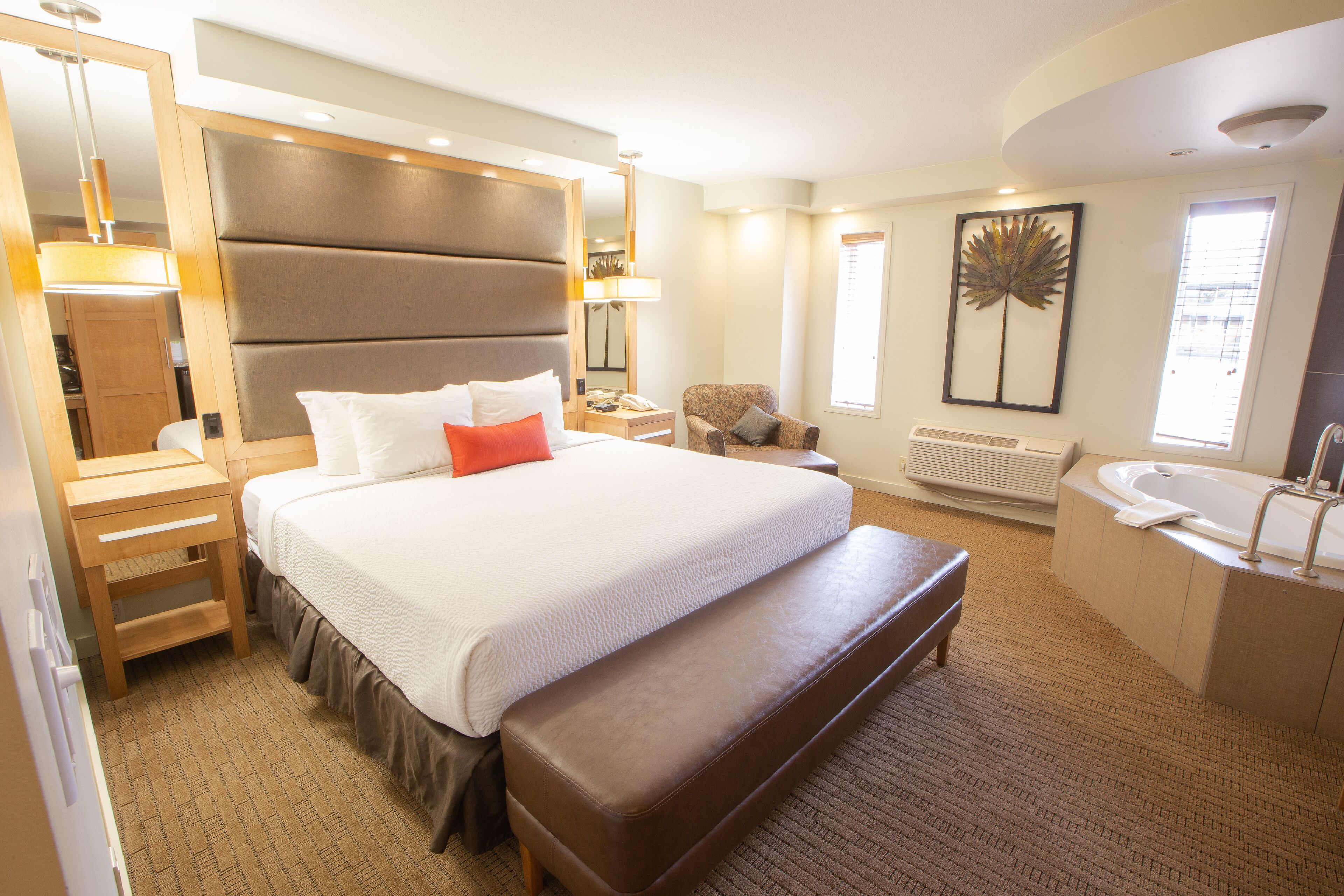 Quarto de hotel com uma cama grande, um banco, uma escrivaninha, uma cadeira, um banheiro com banheira e um espelho amplo.