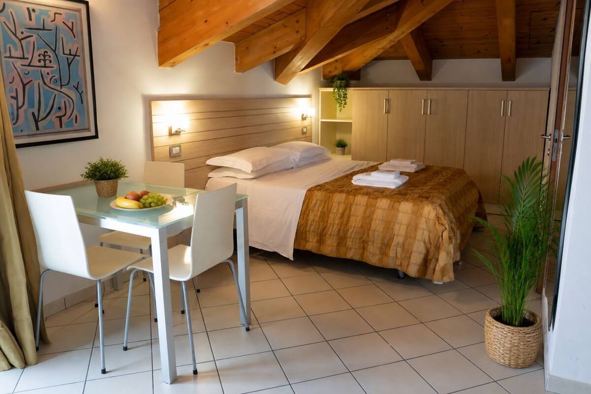Quarto com cama, mesa de cabeceira e escrivaninha com cadeiras. Há um quadro na parede e uma planta no chão.