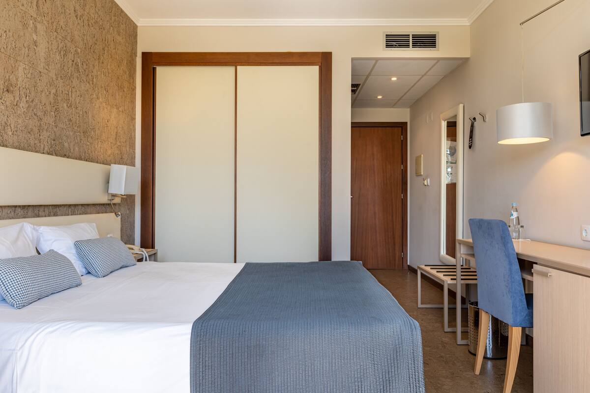Quarto de hotel com cama, mesa de cabeceira, luminária e escrivaninha com cadeira.