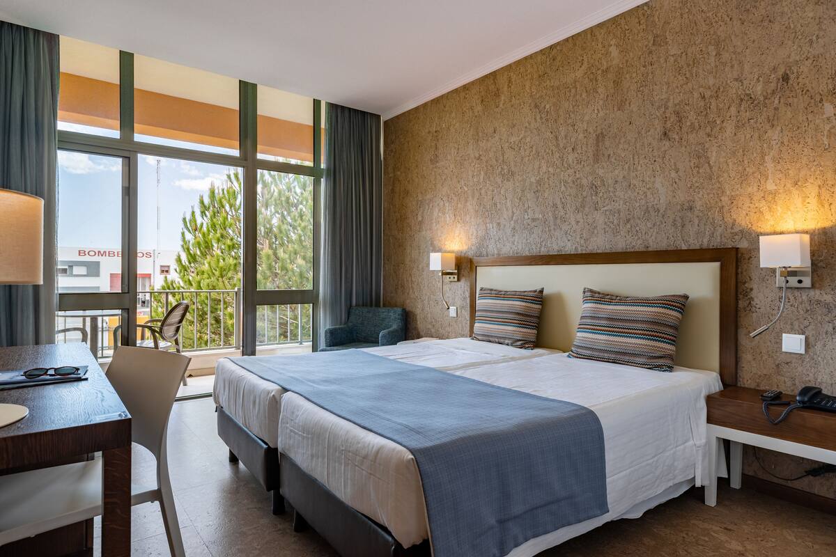Quarto de hotel com uma cama grande, duas mesas de cabeceira, uma escrivaninha com cadeira e vista para o exterior.