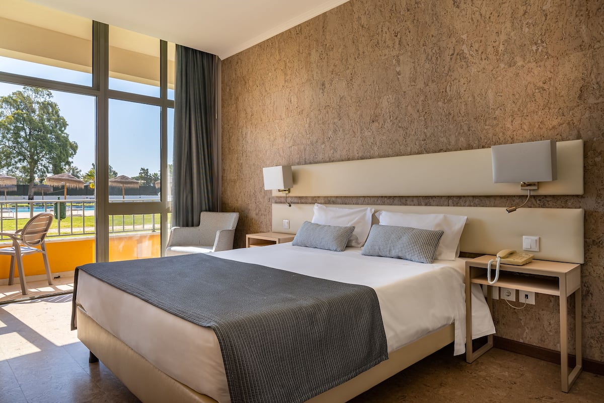 Quarto de hotel moderno com uma cama grande, uma mesa de cabeceira com telefone, e vista para uma piscina e árvores através de grandes janelas.