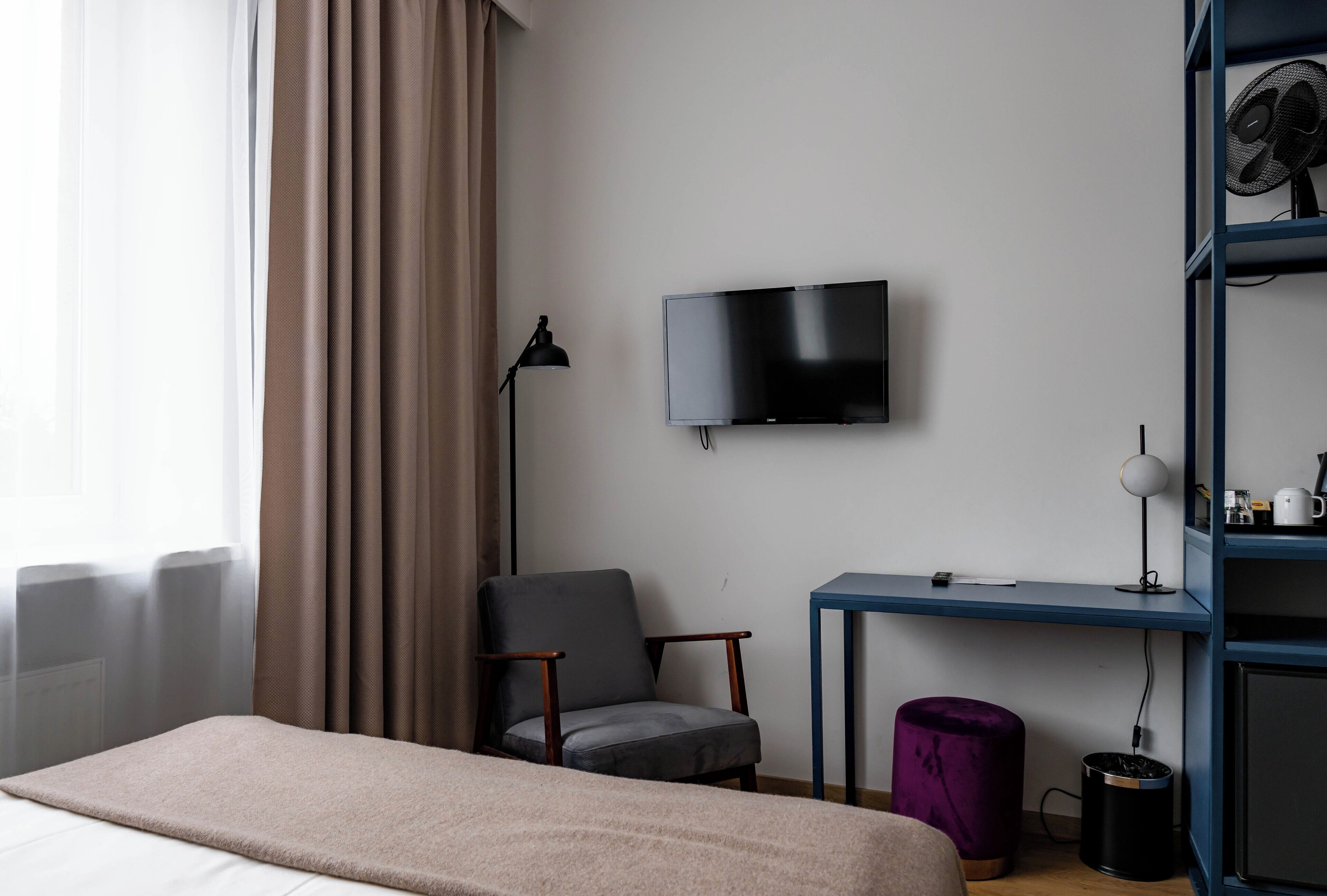 Quarto de hotel com uma cama, uma escrivaninha, uma cadeira, uma televisão, um abajur, um cesto de lixo e um radiador.