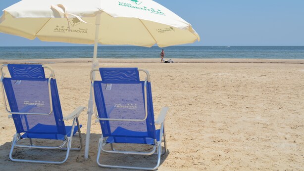 Duas cadeiras de praia azuis sob um guarda-sol branco, com a marca 'Hotel Argentina', numa praia arenosa com o oceano ao fundo.