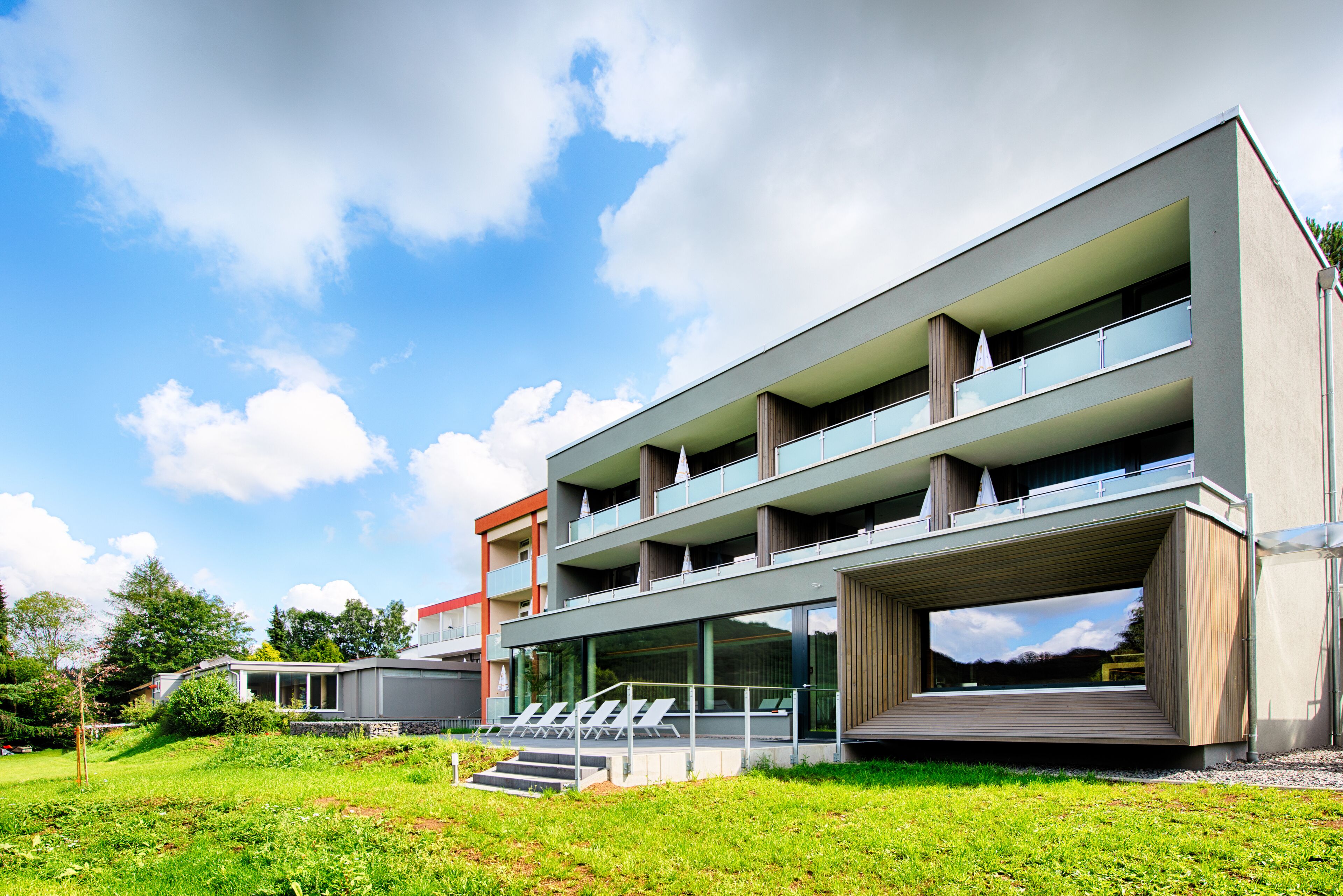 Um edifício moderno de vários andares, com varandas, janelas amplas e uma área gramada na frente.