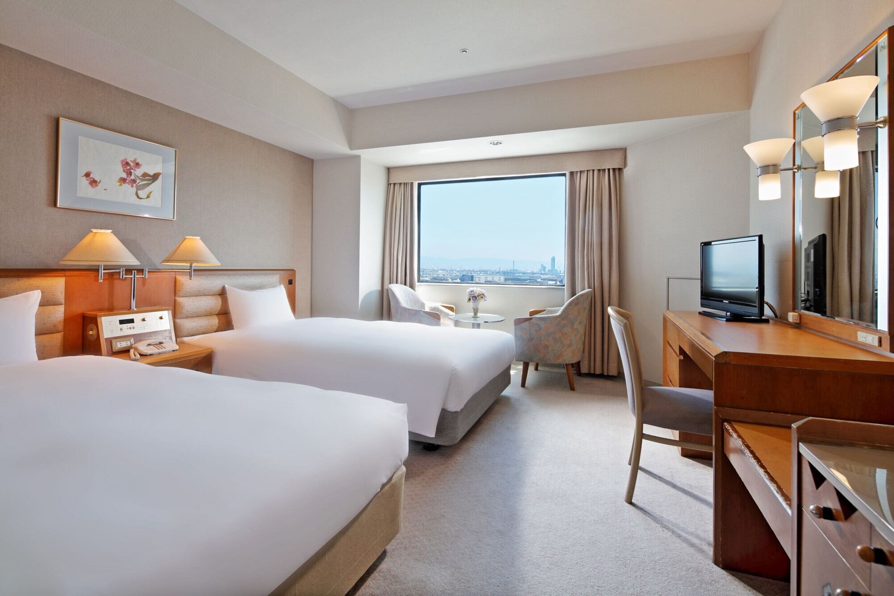 Quarto de hotel com duas camas, uma escrivaninha, uma cadeira, televisão e uma janela grande com vista para a cidade.