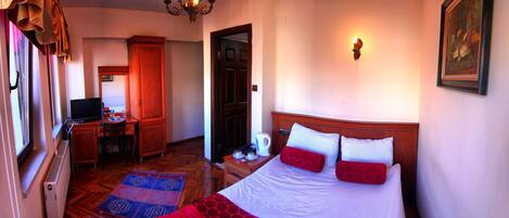 Quarto de hotel com cama, secretária com computador, cadeira, janela com cortinas, radiador, luminária na parede e um quadro.