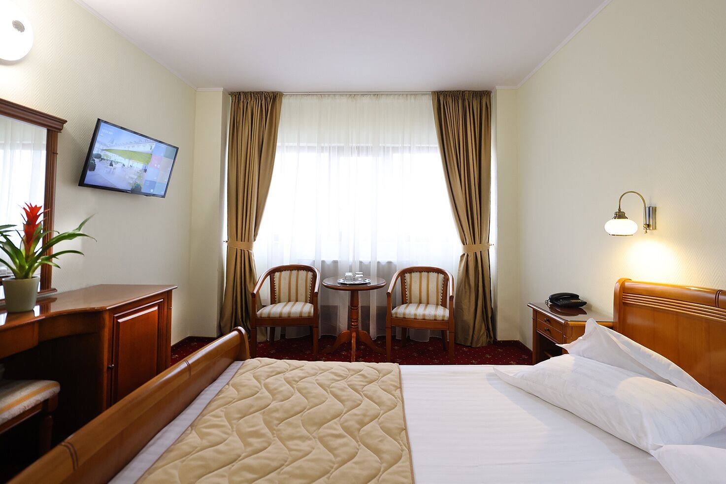 Quarto de hotel com uma cama, duas cadeiras, uma escrivaninha, uma mesinha, uma televisão, cortinas e um abajur.