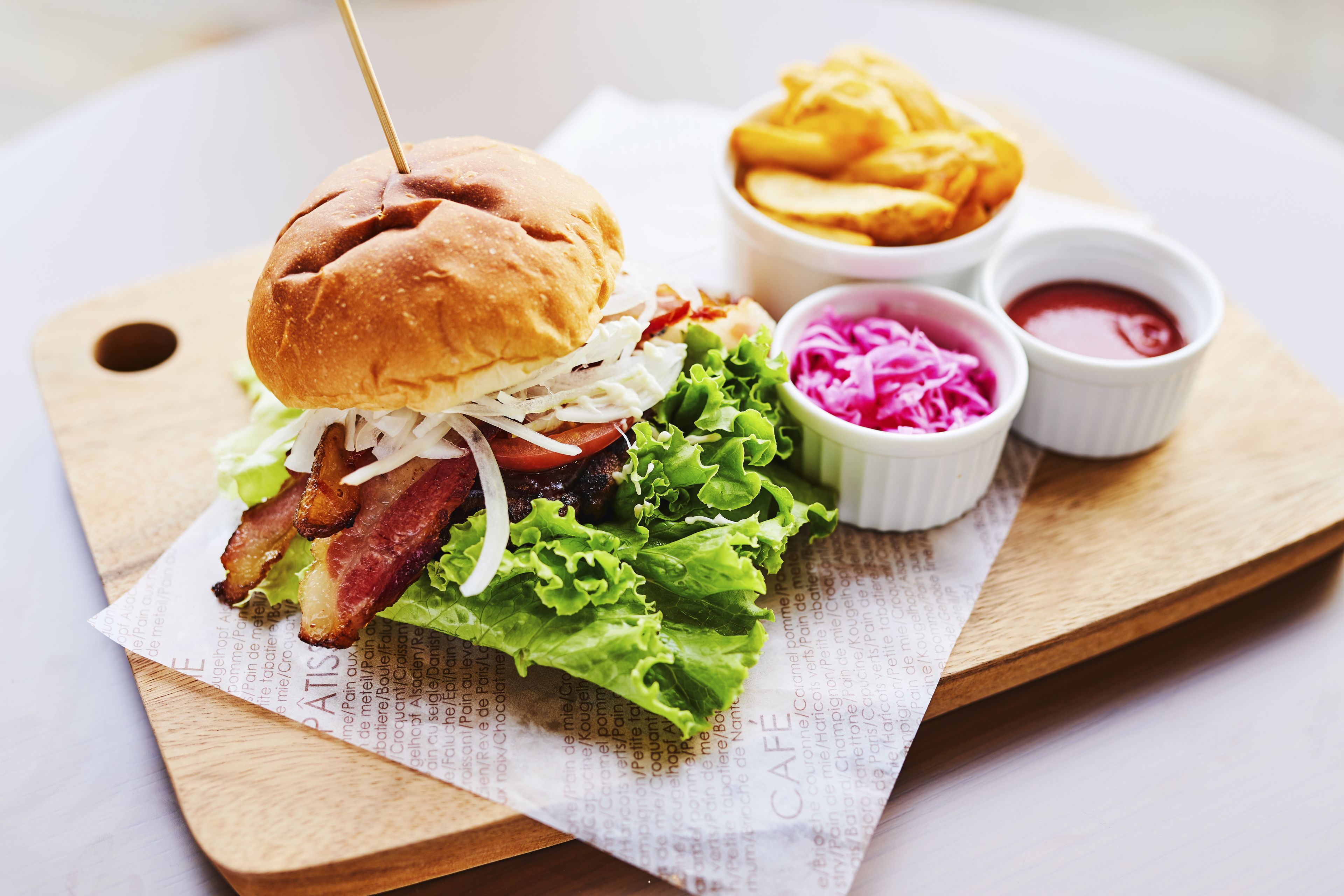 Um hambúrguer com bacon, alface e tomate sobre uma tábua de madeira, acompanhado de batatas fritas e dois molhos.