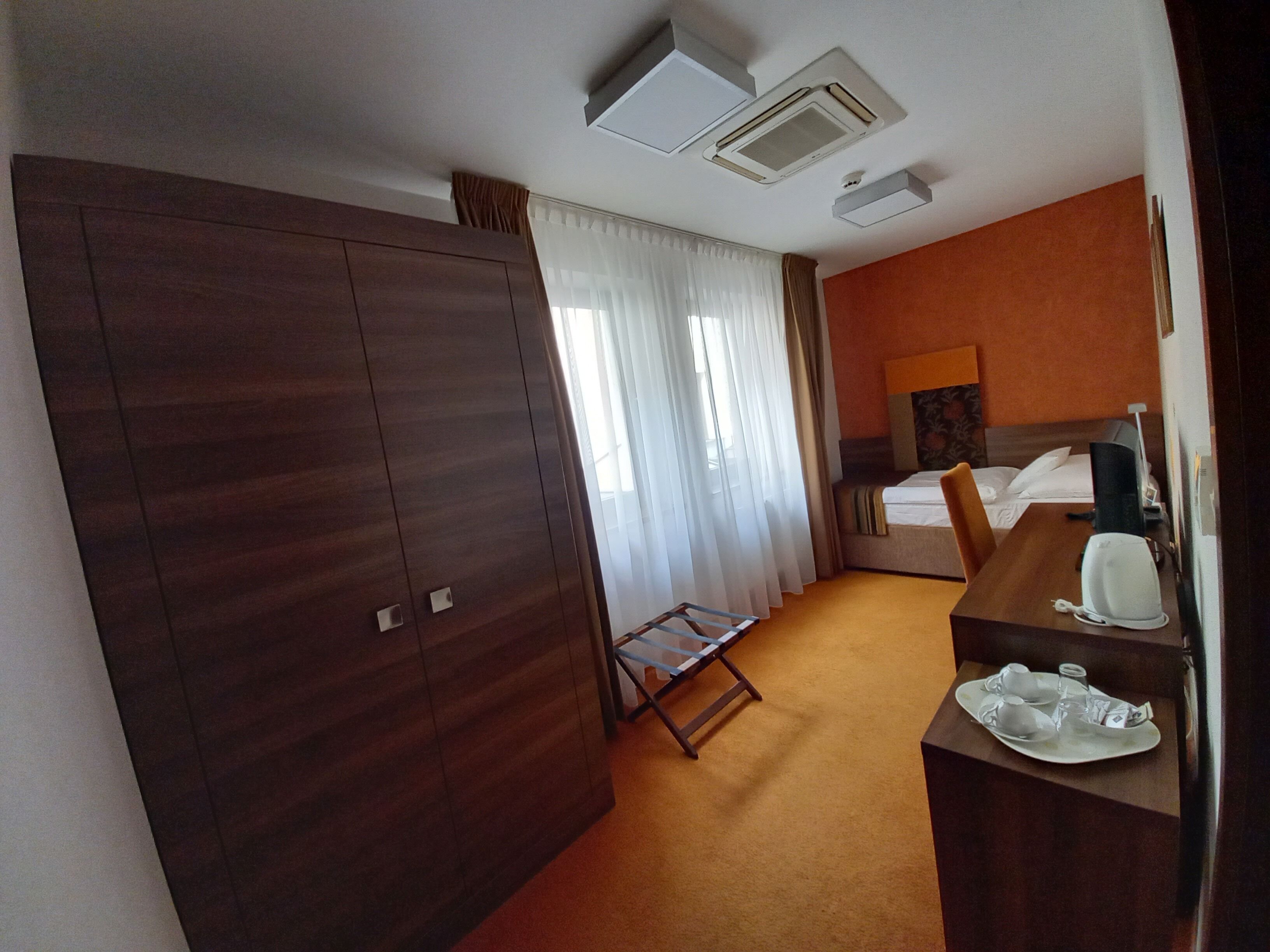 Quarto de hotel com guarda-roupa, secretária com bandeja de chá e chaleira, cama com cabeceira e janela com cortinas.