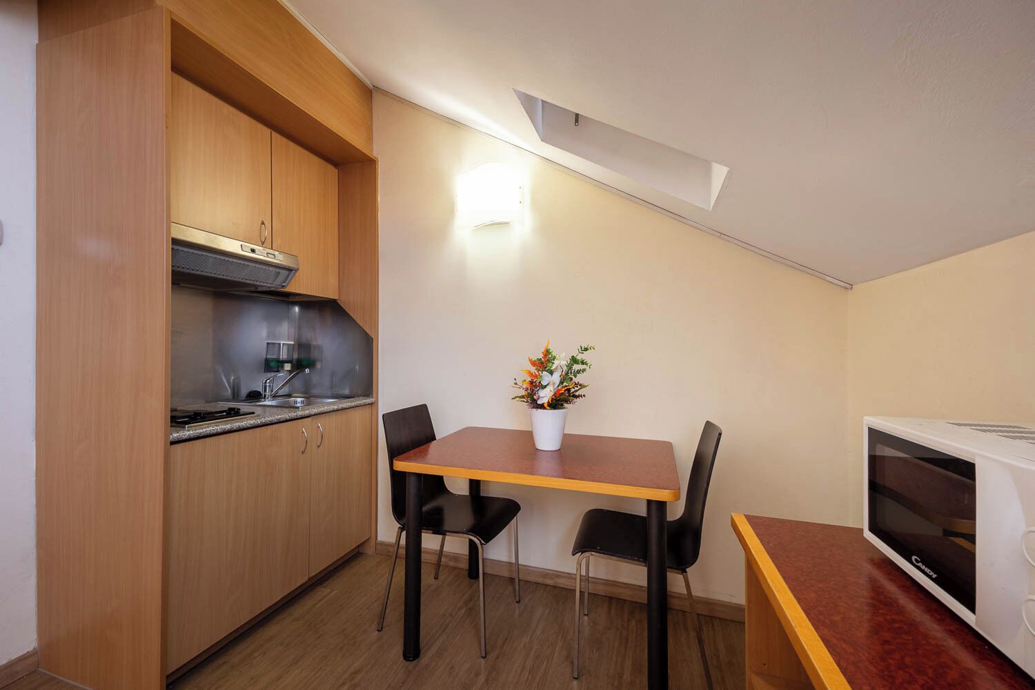 Cozinha compacta com armários de madeira, fogão e micro-ondas. Há uma mesa de jantar com duas cadeiras e um vaso de planta.