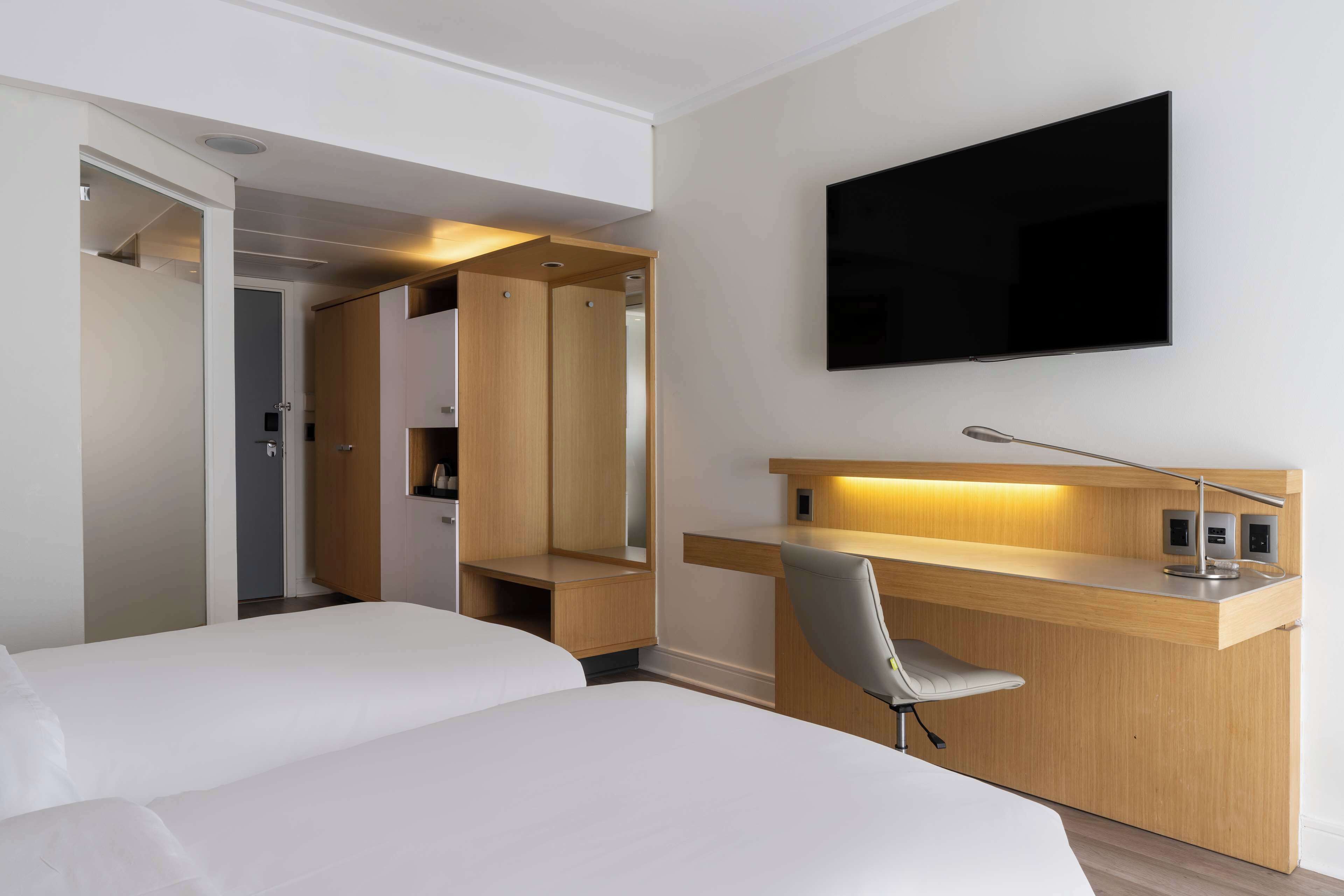 Quarto de hotel moderno com duas camas, uma escrivaninha com cadeira, televisão na parede e guarda-roupa.