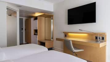 Quarto de hotel moderno com duas camas, uma escrivaninha com cadeira, televisão na parede e guarda-roupa.