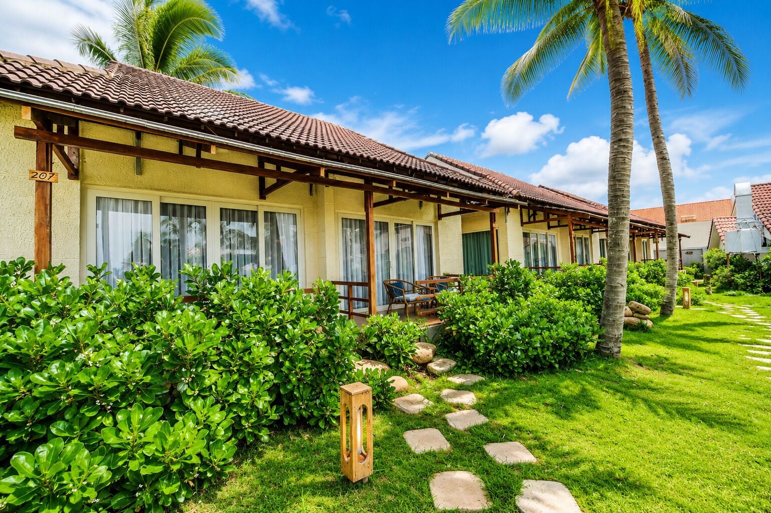 Um hotel com área de estar coberta ao ar livre, um jardim bem cuidado com palmeiras e um céu claro.