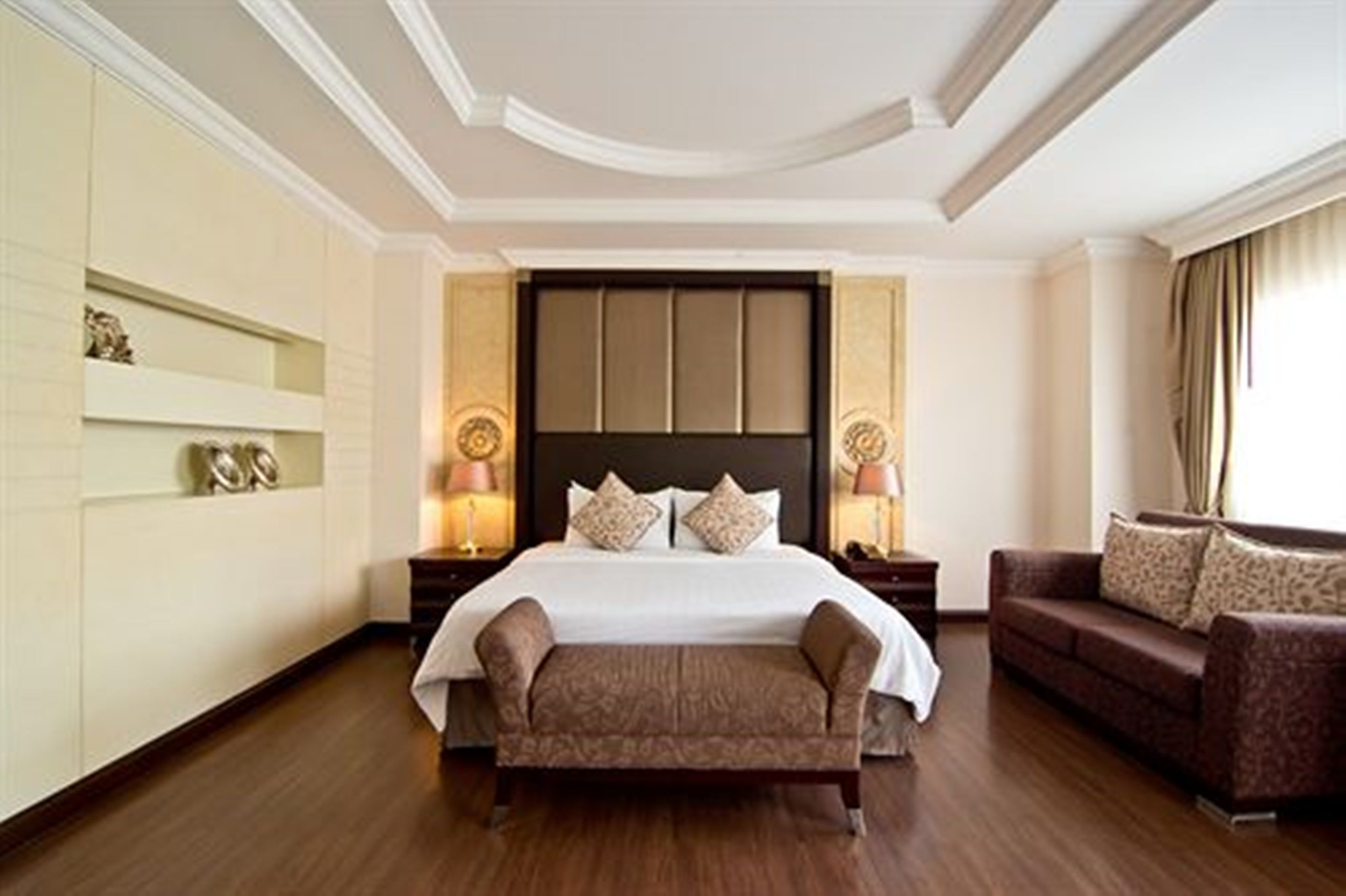 Quarto de hotel com uma cama grande, um sofá, mesa de cabeceira com abajures e prateleira na parede com objetos decorativos.