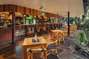 Restaurante com interior em madeira e chão de pedra, com mesas, cadeiras e um espaço de bar com prateleiras de garrafas.