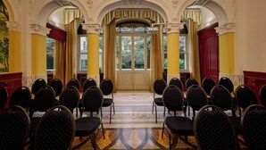 Uma sala de conferências com fileiras de cadeiras, uma janela grande com cortinas e teto decorado.