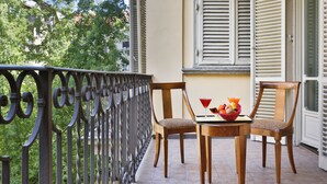 Uma varanda com duas cadeiras de madeira, uma mesa pequena com uma fruteira e uma bebida, e vista para árvores e edifícios.