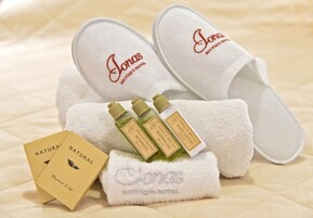 Um par de chinelos brancos com a inscrição "Jonas Boutique Hotel" bordada, uma toalha branca e dois frascos de touca de banho e loção corporal "Natura Natural".