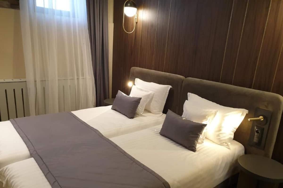 Cama de casal com cabeceira cinza, roupa de cama branca e mesa de cabeceira com abajur.