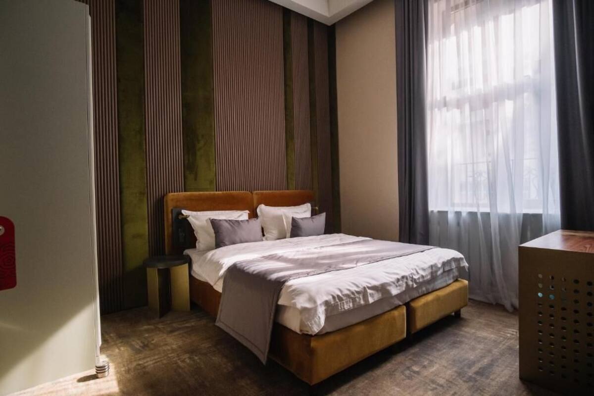 Quarto de hotel com cama, mesas de cabeceira, janela com cortinas e piso de madeira.