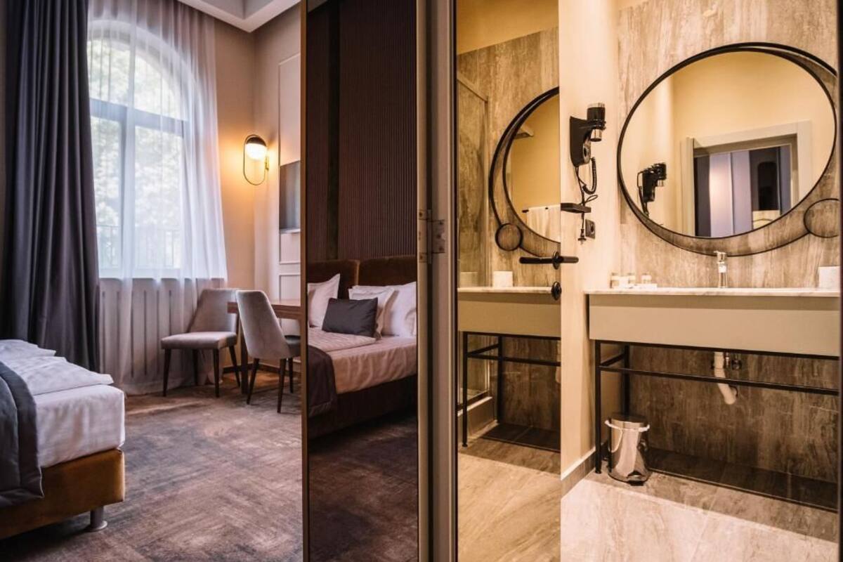 Quarto de hotel com cama, cadeira, escrivaninha, espelho grande e banheiro com pia e espelho.