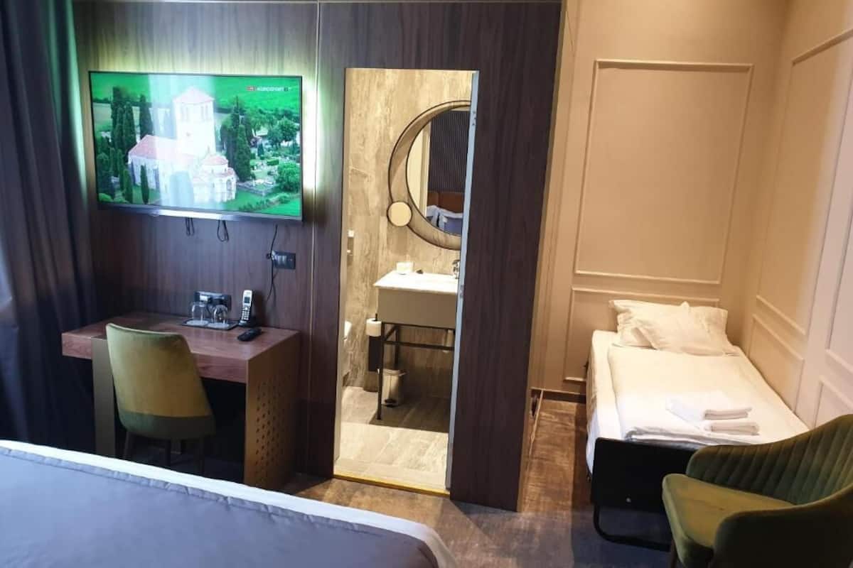 Quarto de hotel com cama, escrivaninha com telefone, cadeira, televisão e banheiro visível através de uma porta aberta.