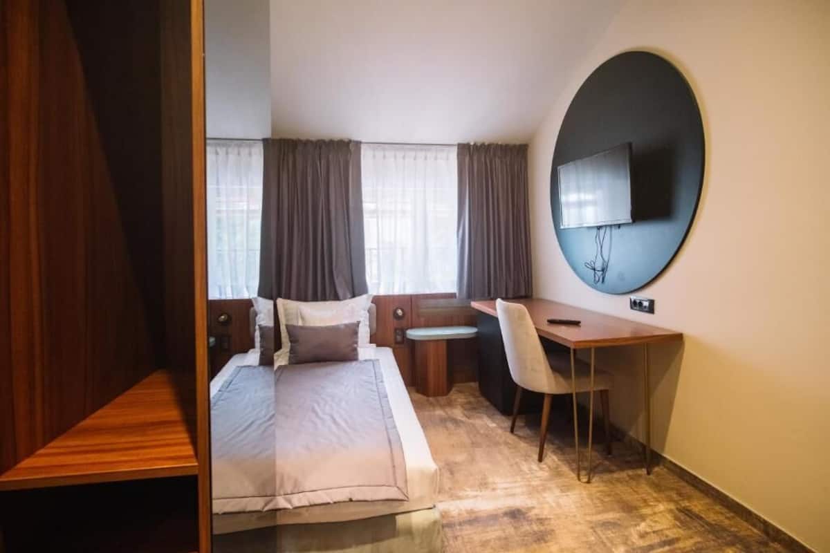 Quarto de hotel com cama, escrivaninha, cadeira e televisão fixada na parede.