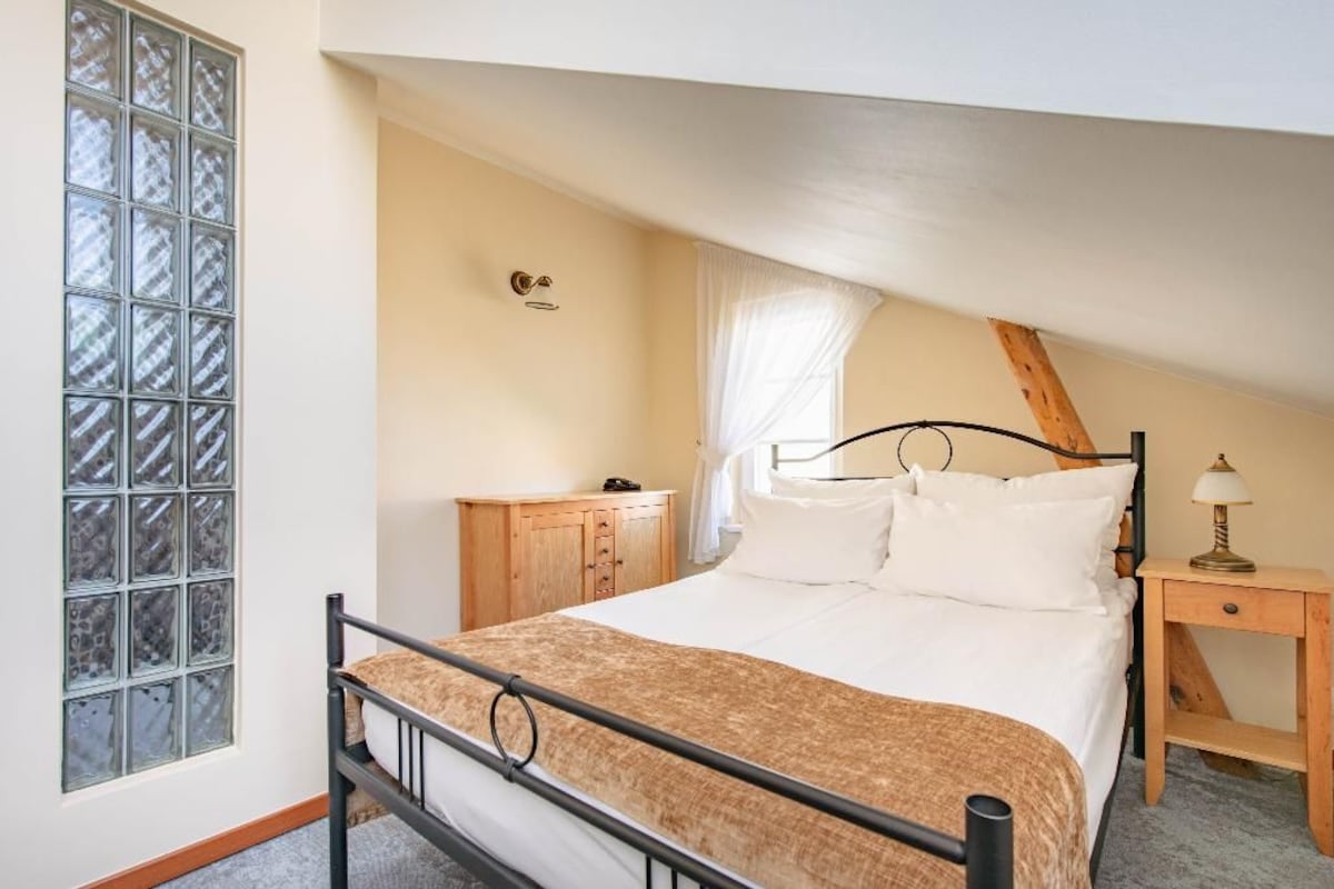 Quarto com cama, mesa de cabeceira, cômoda de madeira e janela com vista.
