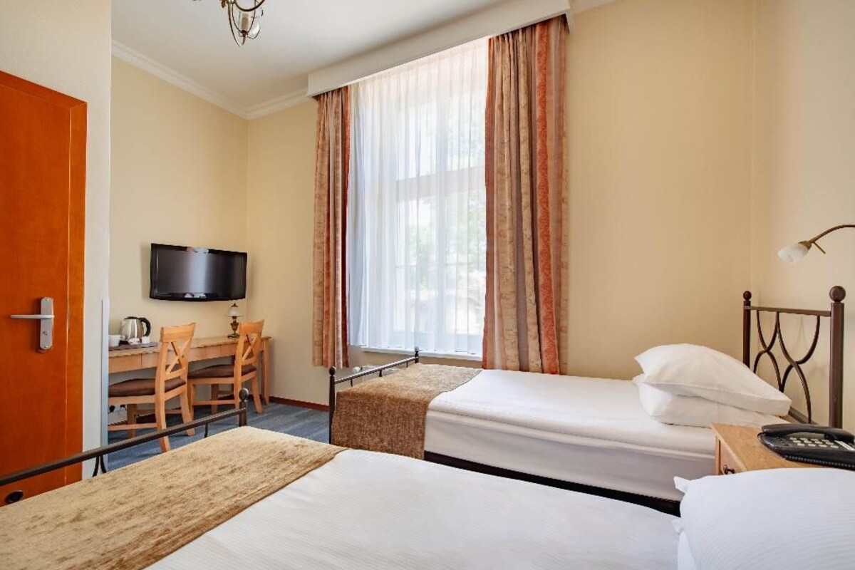 Quarto de hotel com duas camas, televisão, escrivaninha com cadeiras e janela com cortinas.