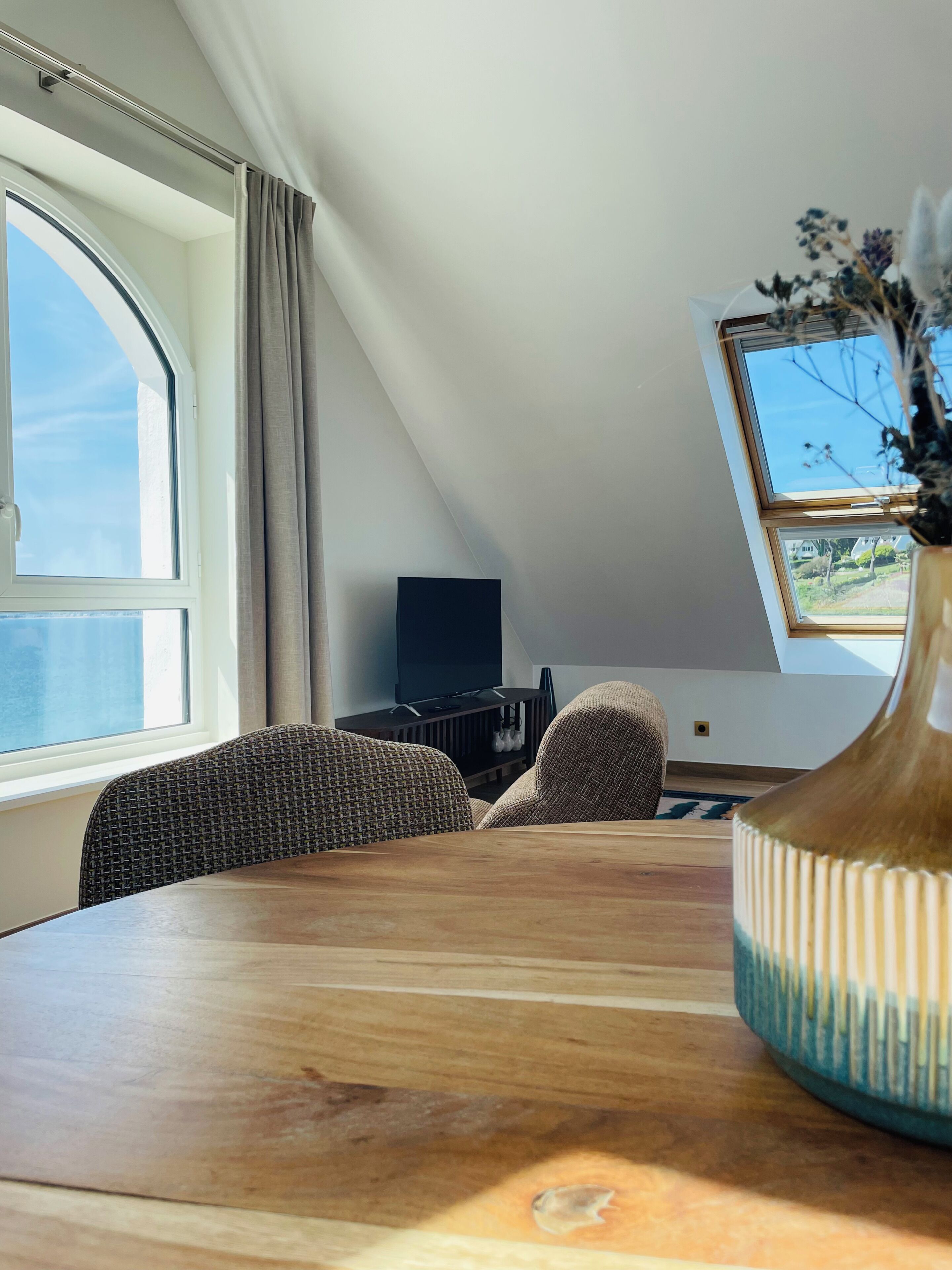Um quarto com uma mesa redonda de madeira, duas cadeiras estampadas, uma televisão sobre um suporte e uma janela com cortinas.