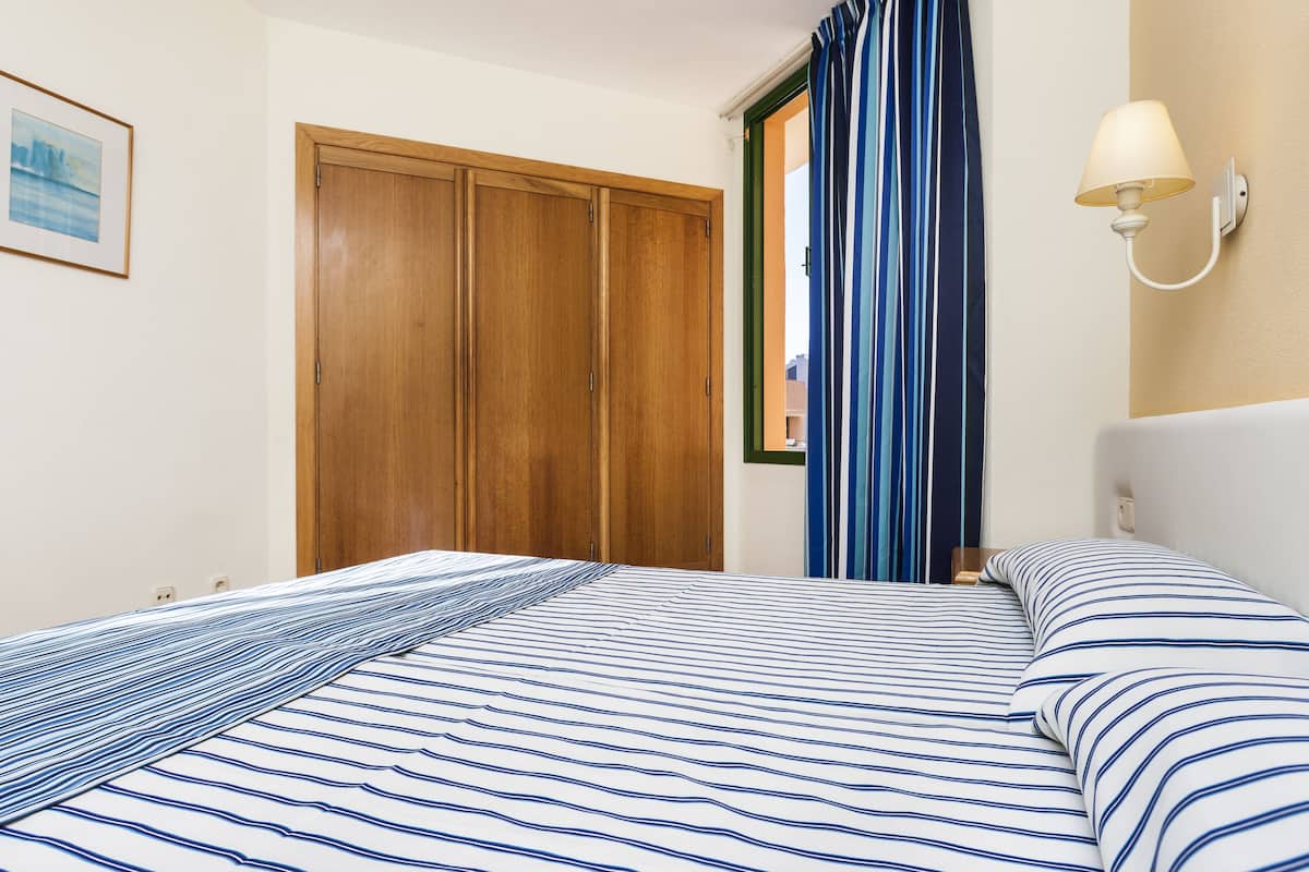 Quarto com cama, guarda-roupa de madeira, cortina listrada e luminária fixa na parede.