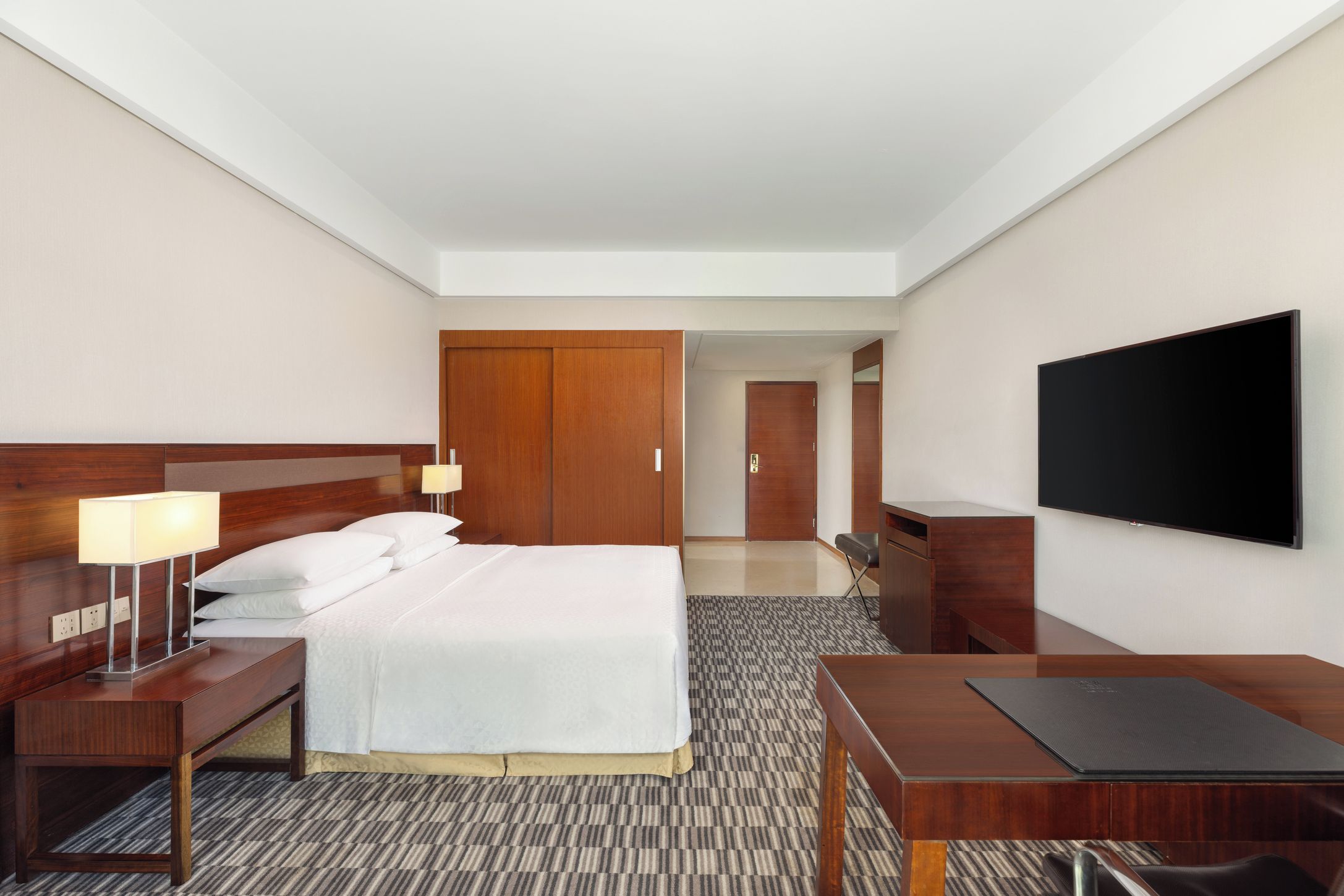Quarto de hotel com cama, mesas de cabeceira, escrivaninha, TV de tela plana e armário.