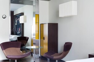 Quarto de hotel moderno com uma mesa de vidro, duas poltronas estofadas, uma cama e um guarda-roupa.