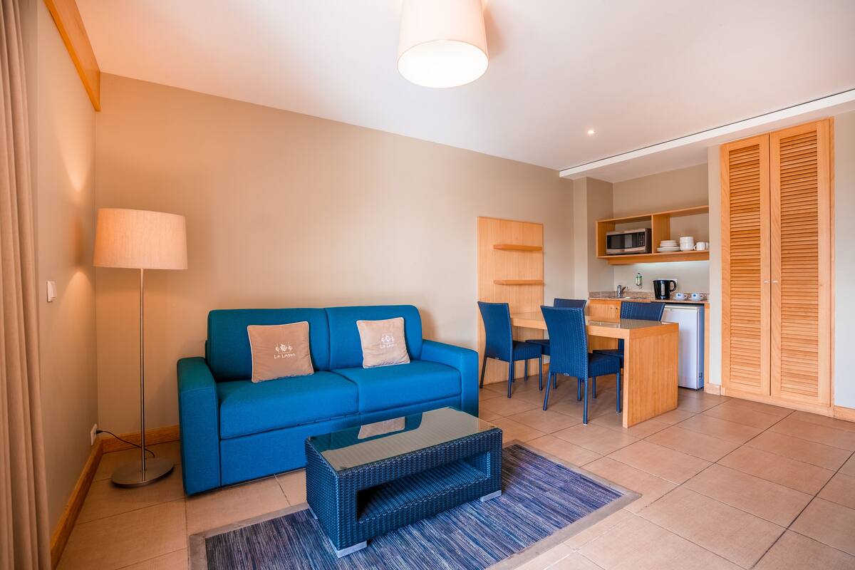 Uma sala de estar moderna com um sofá azul, uma mesa de centro e um espaço de jantar com cadeiras, além de uma pequena cozinha ao fundo.