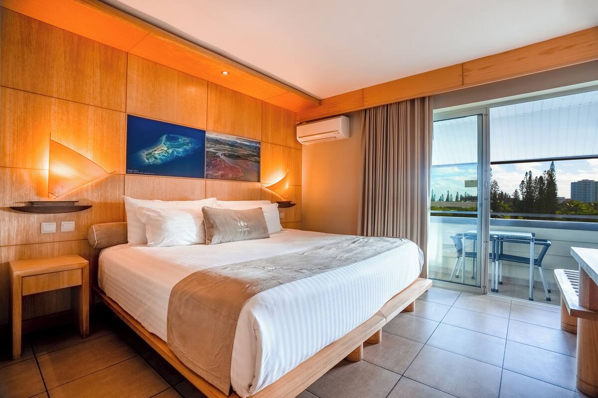 Quarto de hotel moderno com uma cama grande, painéis de madeira, varanda com mesa e cadeiras, e vista para a cidade.
