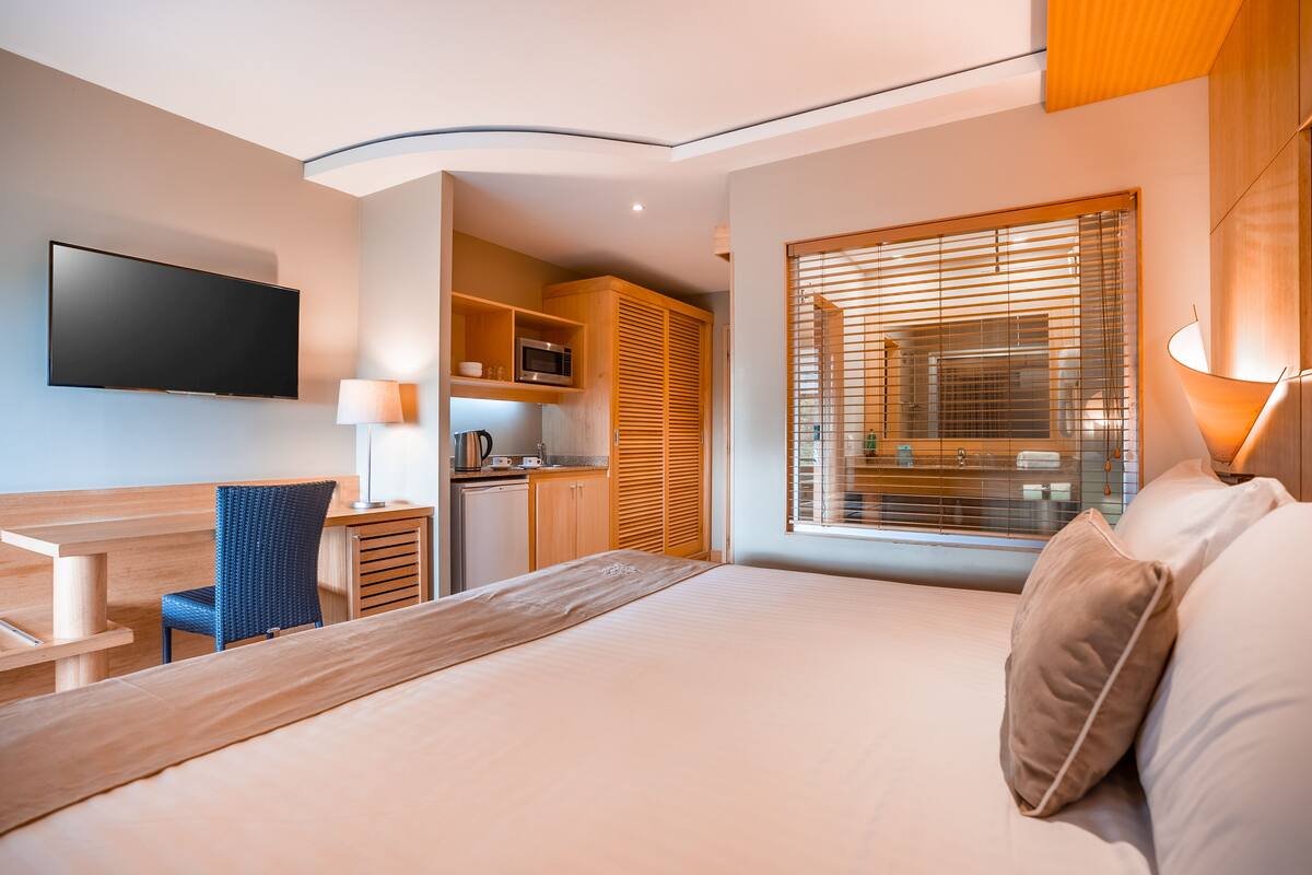 Quarto de hotel moderno com televisão de ecrã plano, secretária com cadeira, cama com almofadas, kitchenette e uma janela ampla com persianas.