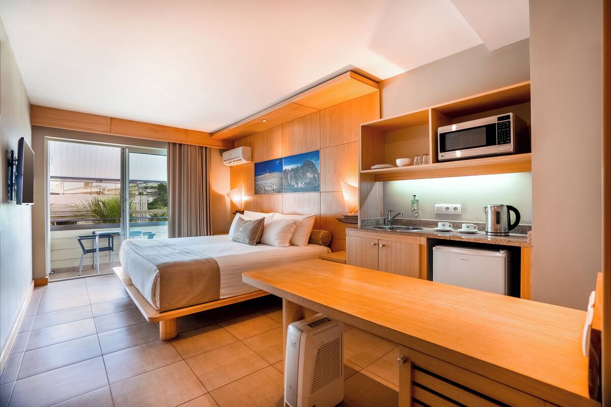 Quarto de hotel moderno com cama, cozinha compacta e varanda com vista.