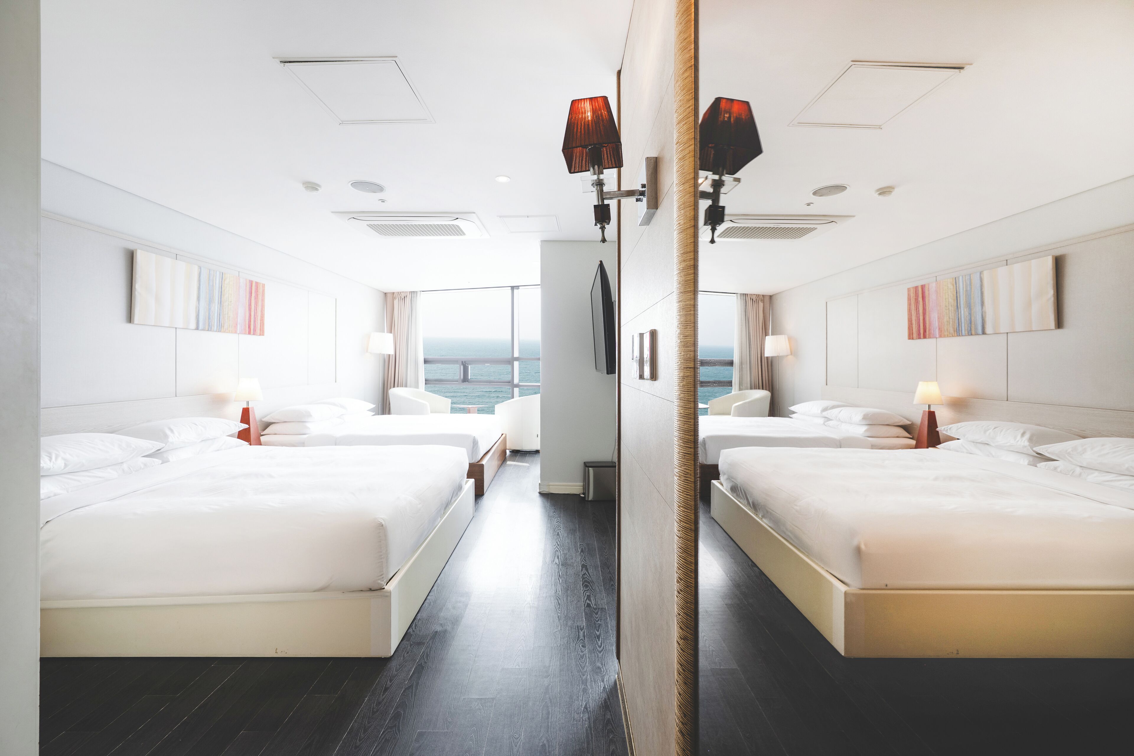 Quarto de hotel moderno com duas camas, um espelho grande e vista para o mar.