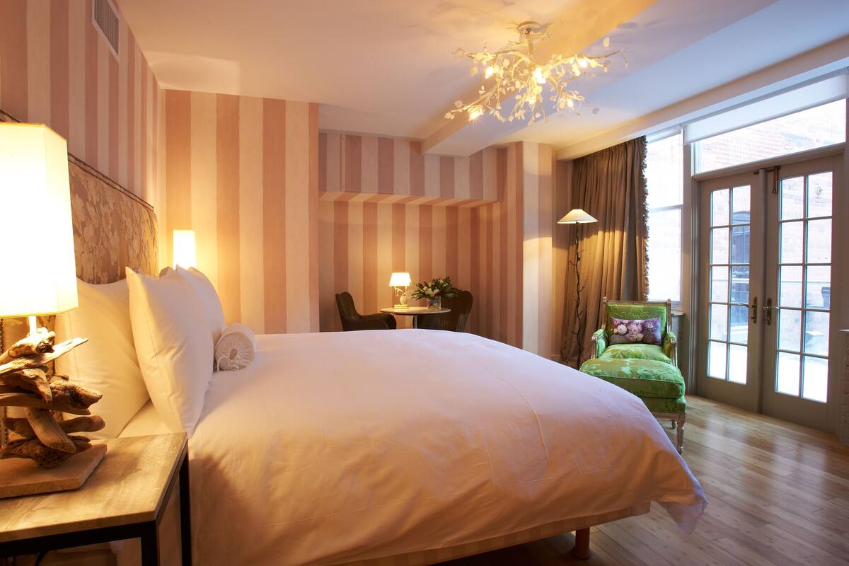 Quarto de hotel com uma cama grande, uma mesa pequena com abajur, uma poltrona verde e vista para o exterior por uma janela ampla.
