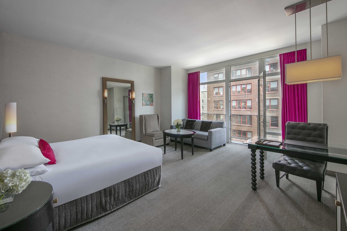 Quarto de hotel com uma cama grande, um sofá, uma escrivaninha e uma cadeira. Há uma janela com cortinas, um espelho e um abajur.