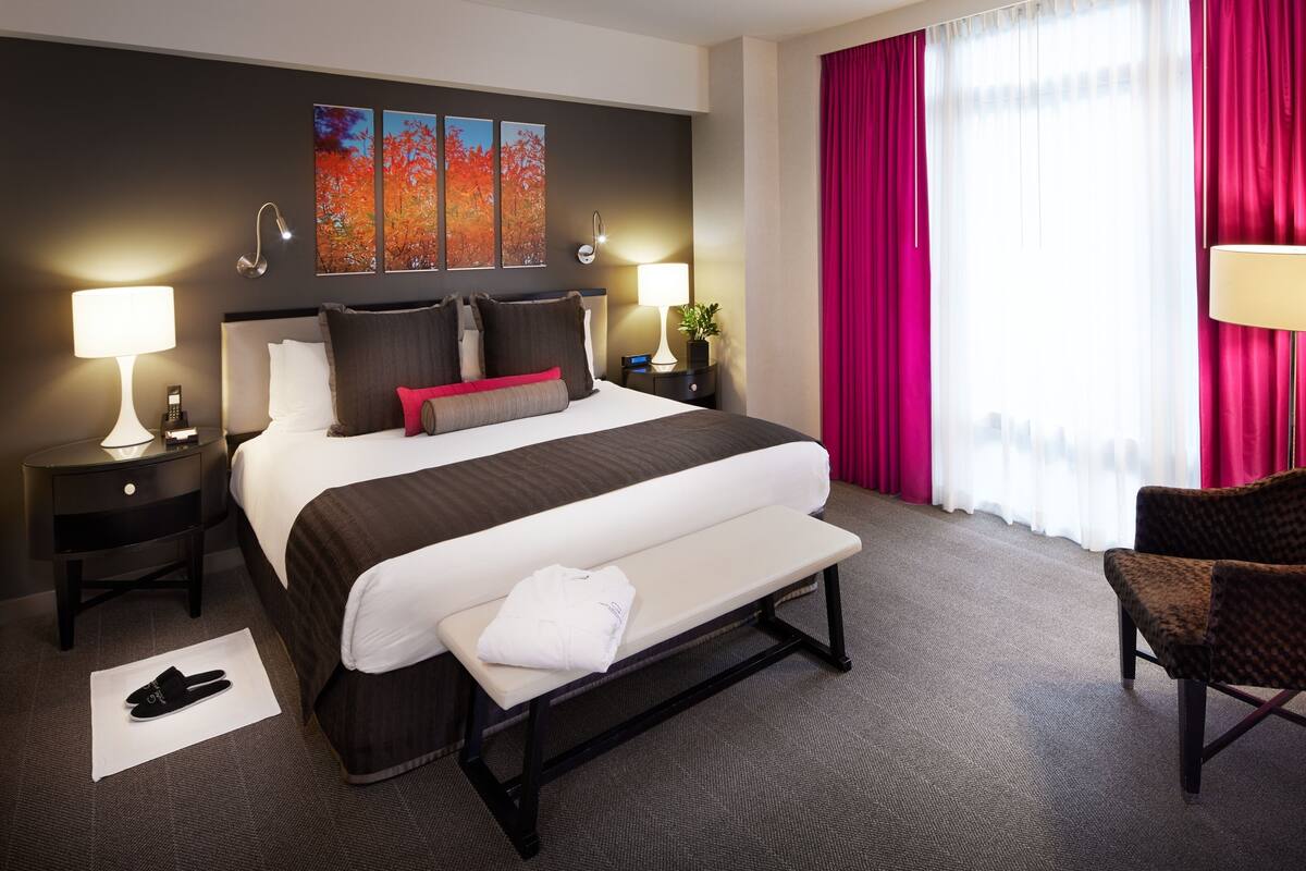 Quarto de hotel com uma cama grande, duas mesas de cabeceira com luminárias, um banco, uma cadeira e uma janela com cortinas.