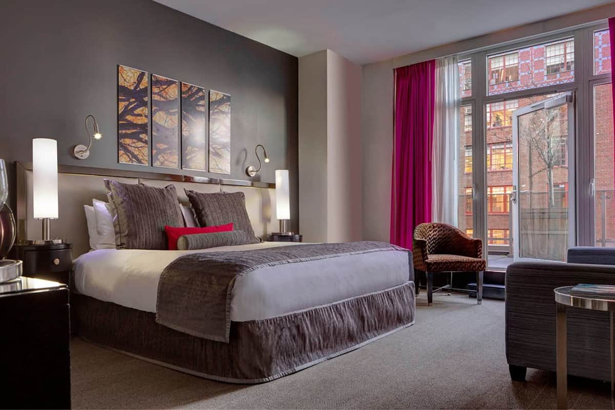 Quarto de hotel moderno com uma cama grande, uma cadeira, uma mesinha e uma janela com cortinas.